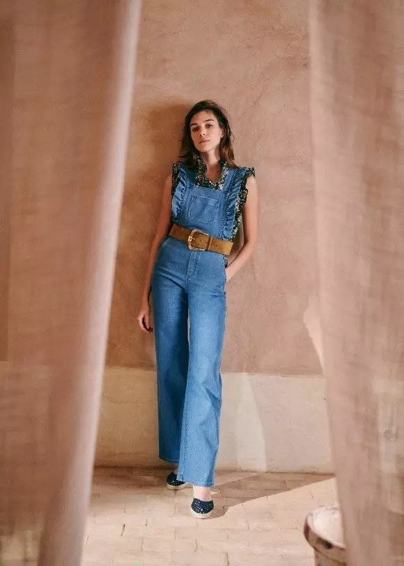 High-waisted denim jumpsuit haum rau ntawm lub duav nrog lub hnab ris pem hauv ntej thiab ruffles ntawm cov hlua. Dav, ncaj ceg.