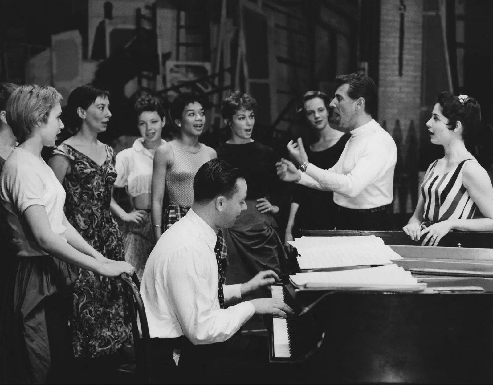 De Leonard Bernstein mat Membere vu West Side Story a Stephen Sondheim um Piano an enger Foto vun 1957.